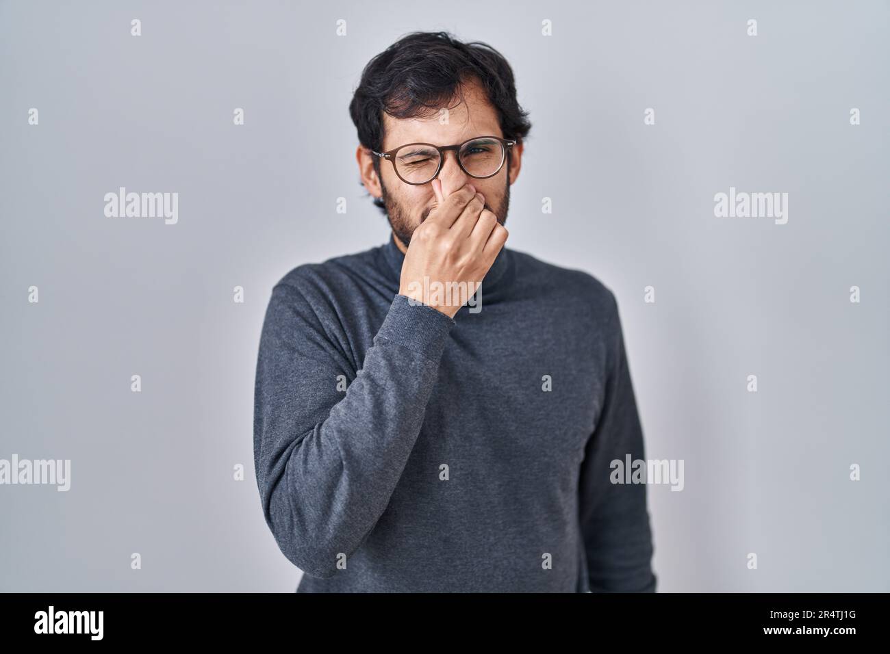 Handsome latin man standing over isolated background smelling something ...
