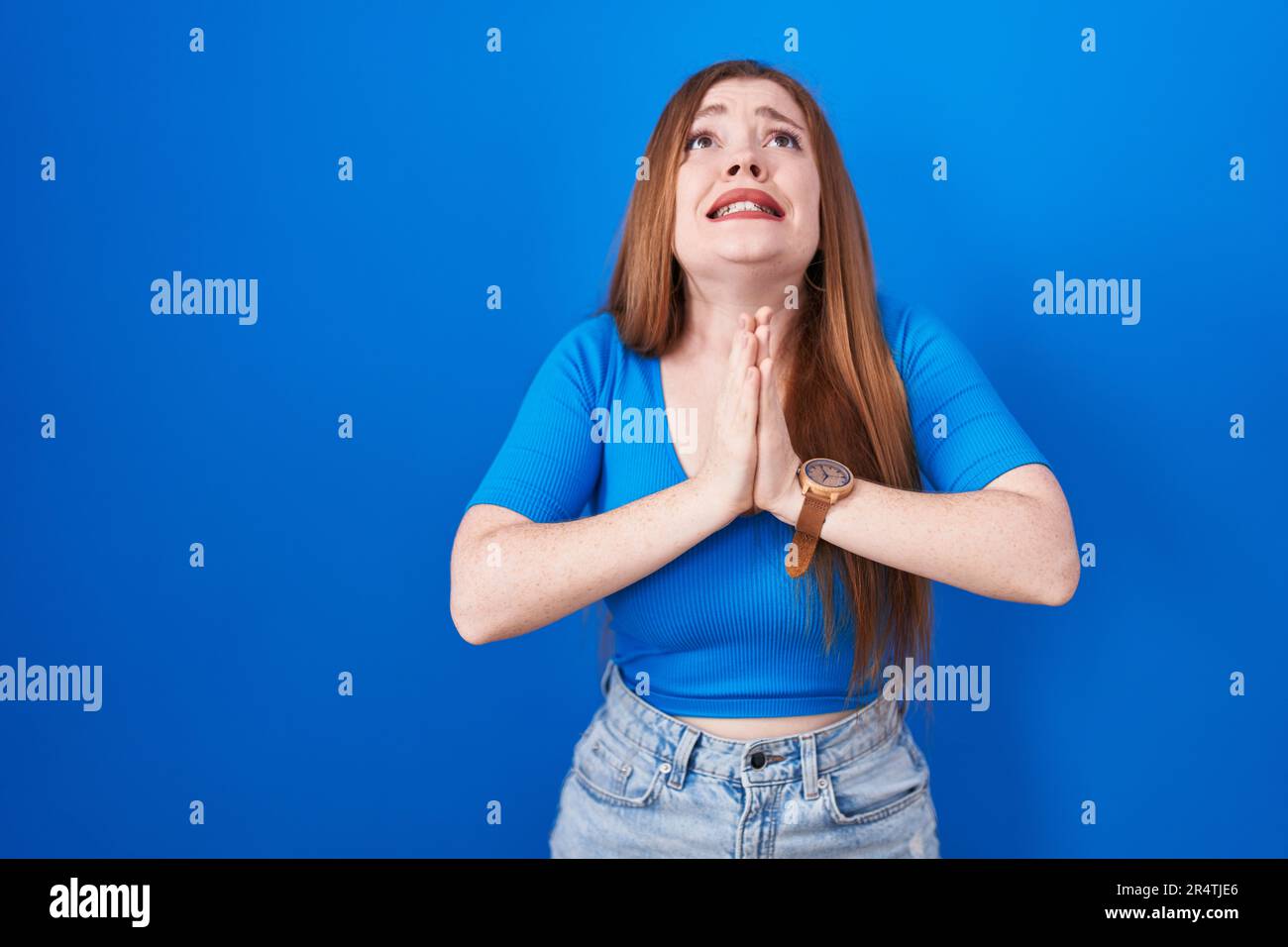 Redhead woman standing over blue background begging and praying with ...