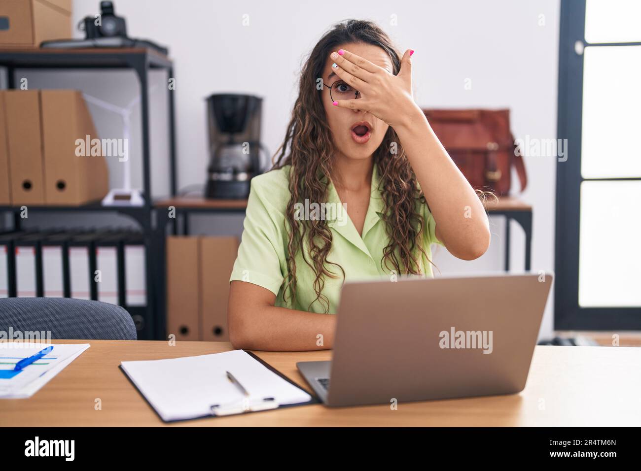 Young hispanic woman working at the office wearing glasses peeking in ...