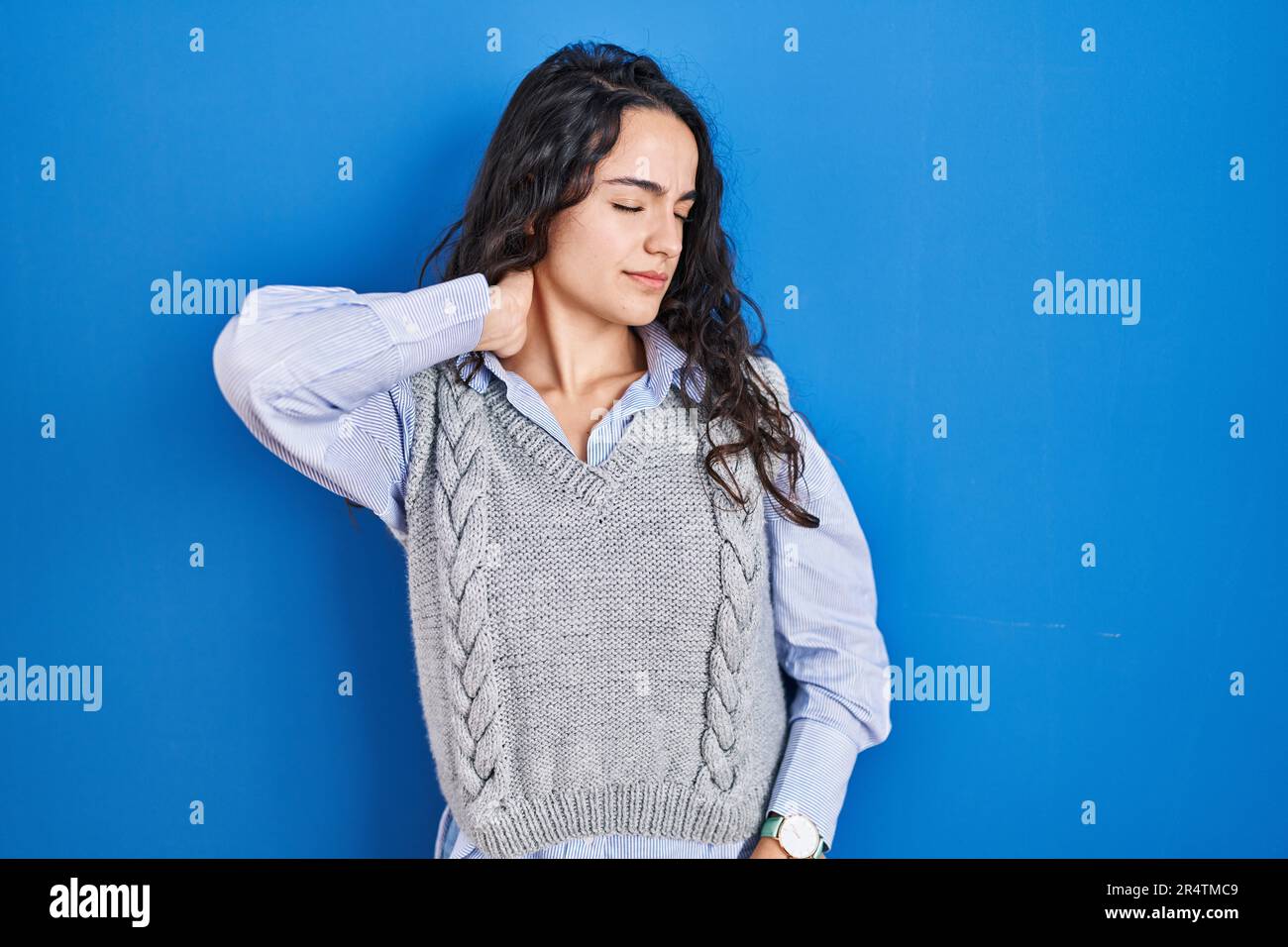 Young brunette woman standing over blue background suffering of neck ...