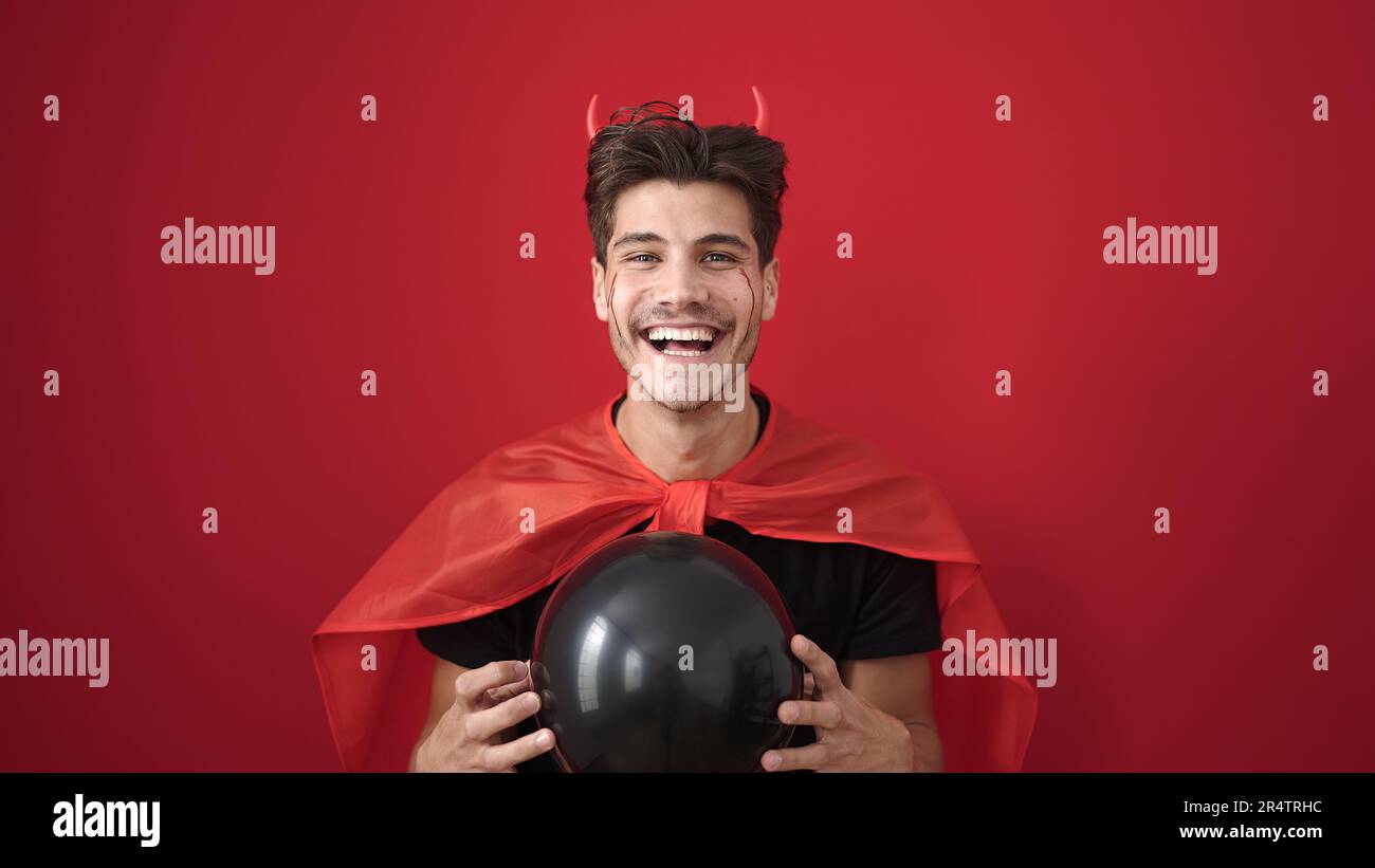 Young hispanic man wearing devil costume holding balloon over isolated ...