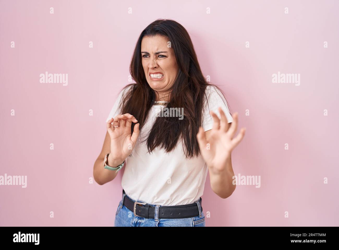 Young brunette woman standing over pink background disgusted expression ...