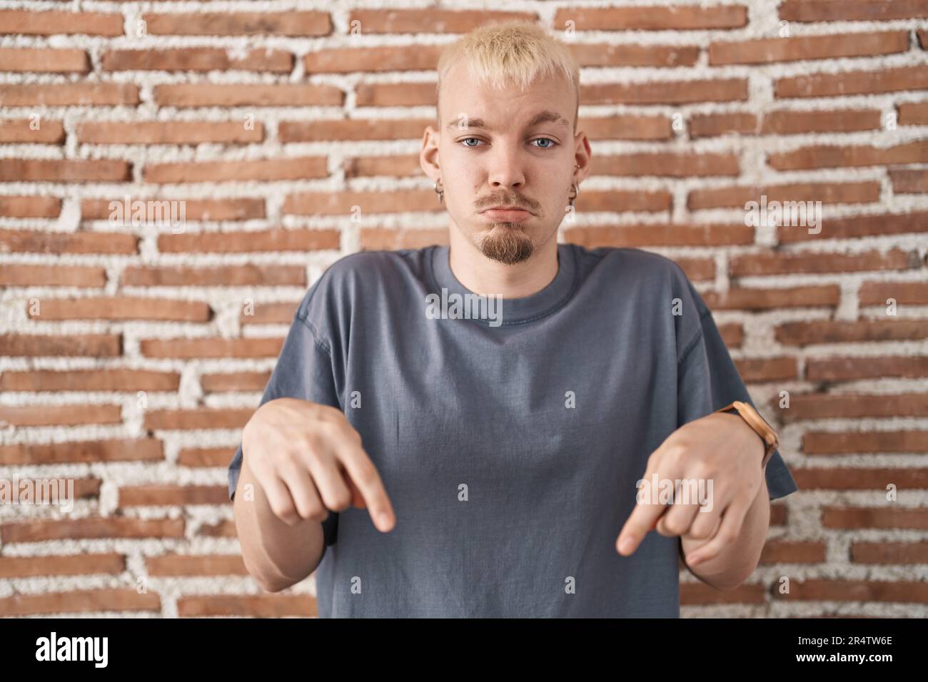 Young caucasian man standing over bricks wall pointing down looking sad ...