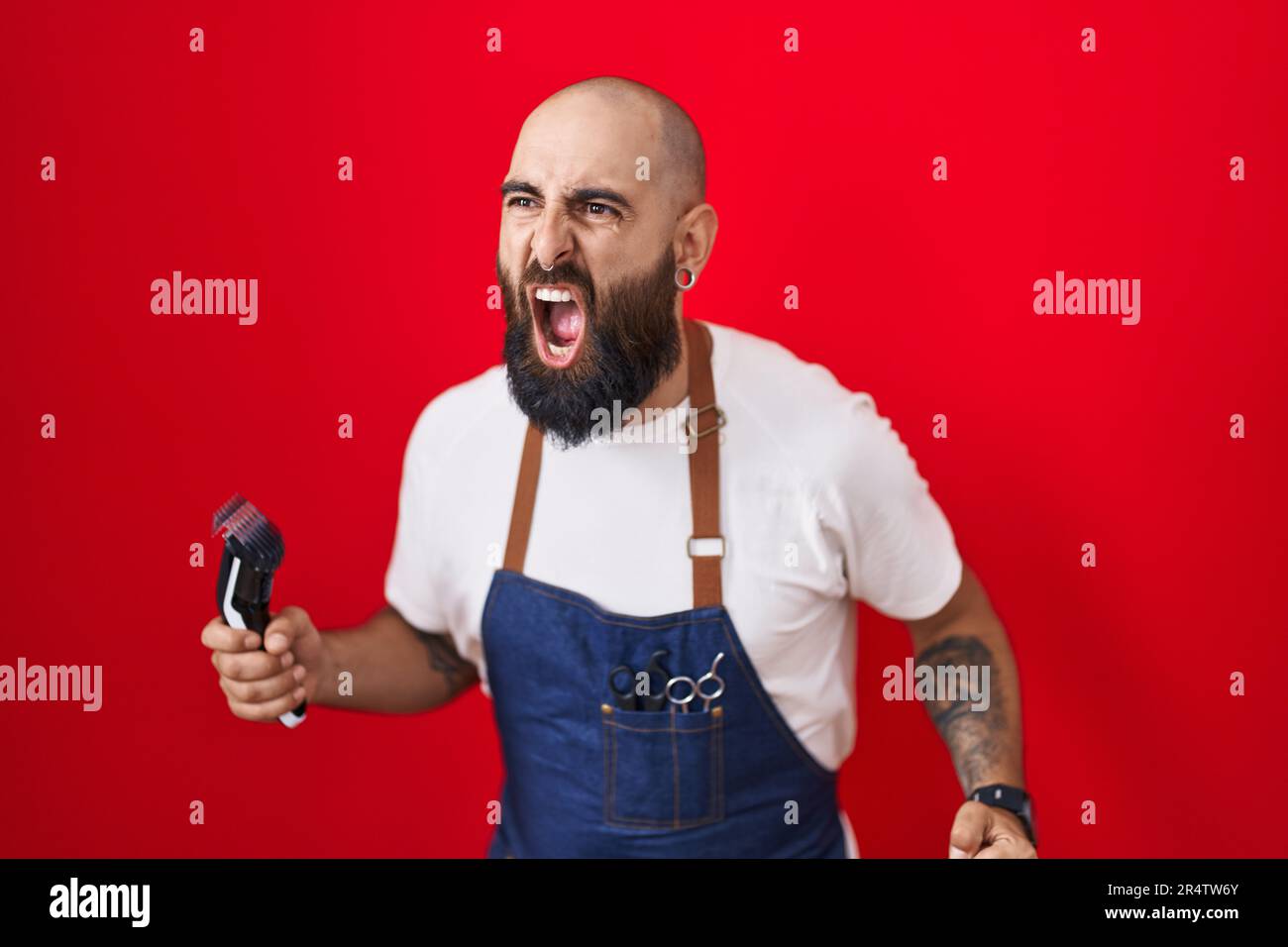 Young hispanic man with beard and tattoos wearing barber apron holding ...