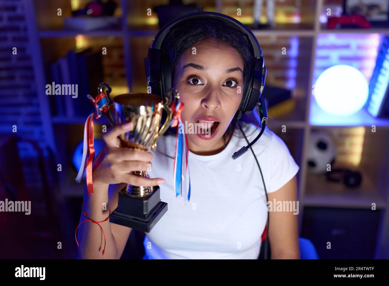 Young arab woman playing video games holding trophy scared and amazed ...