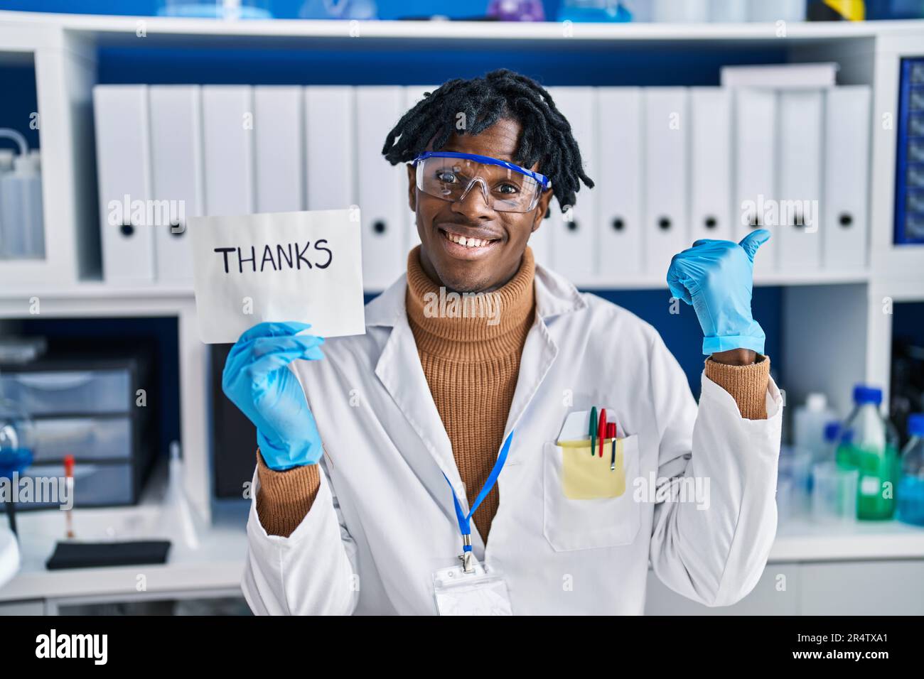 Young african man with dreadlocks working at scientist laboratory pointing thumb up to the side ...