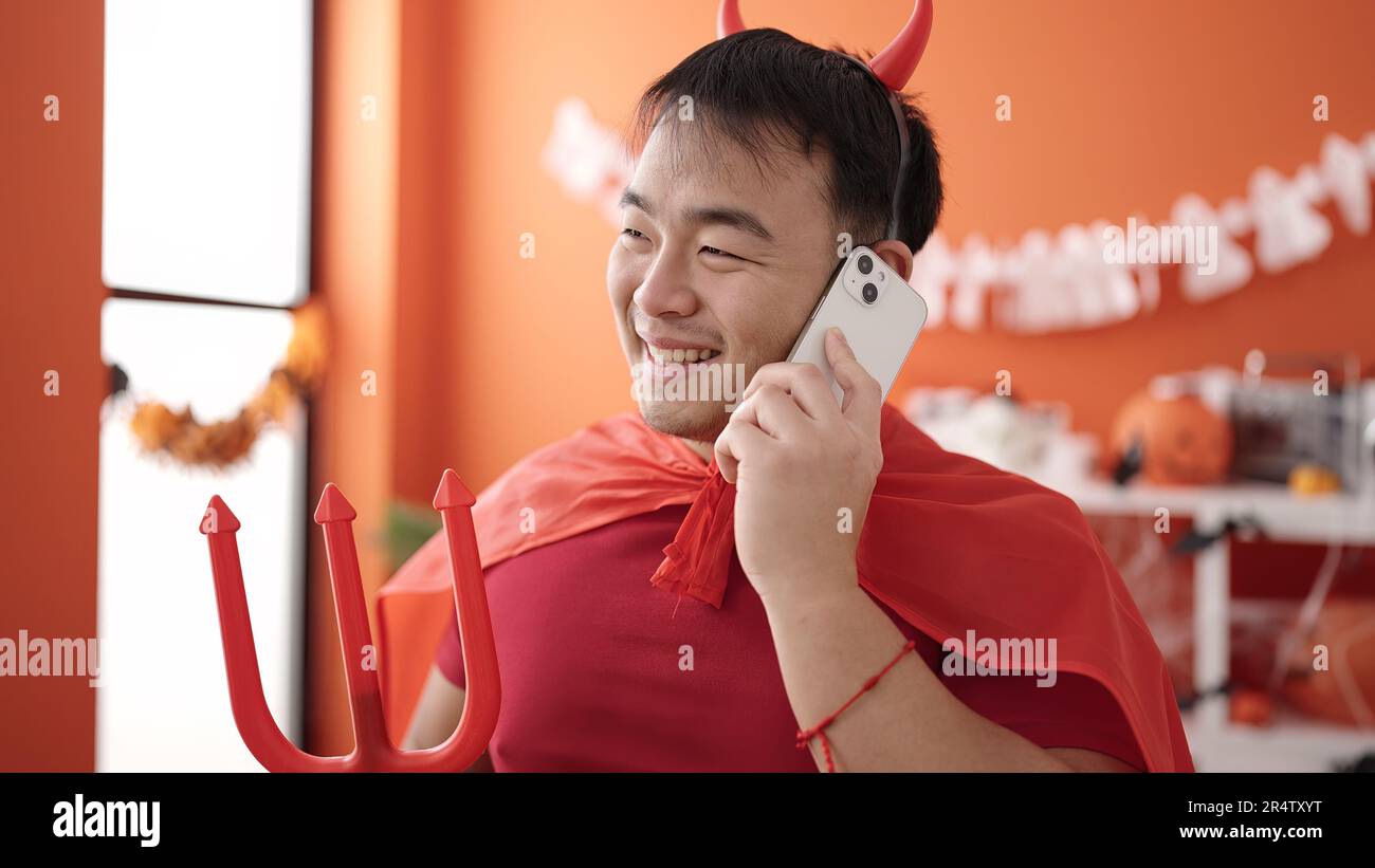 Young chinese man wearing devil costume talking on smartphone at home ...