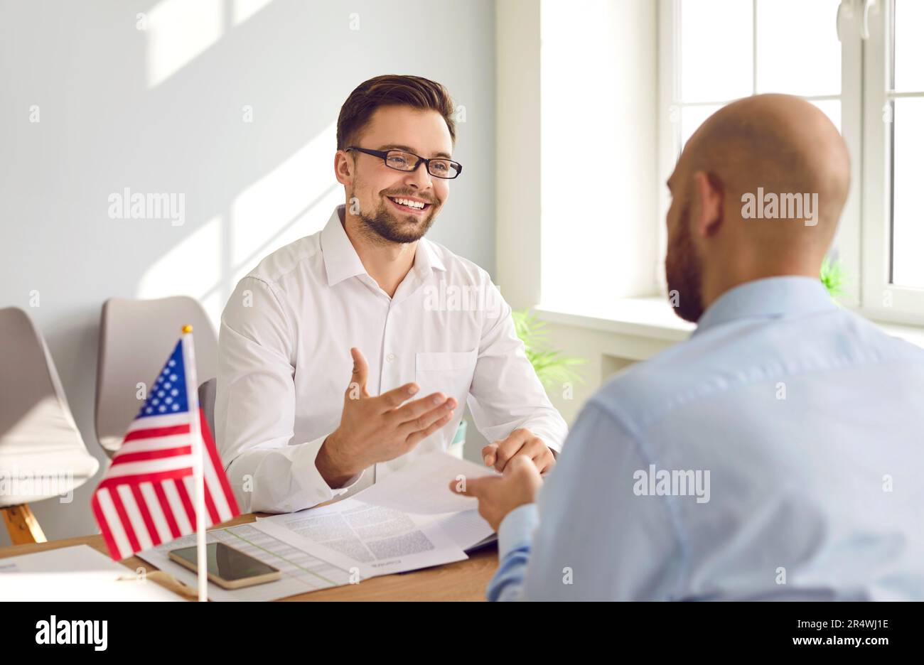 Smiling consul at office desk with American flag talking to man about ...