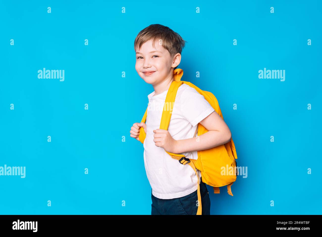 Schoolboy with yellow backpack stands sideways against blue background ...
