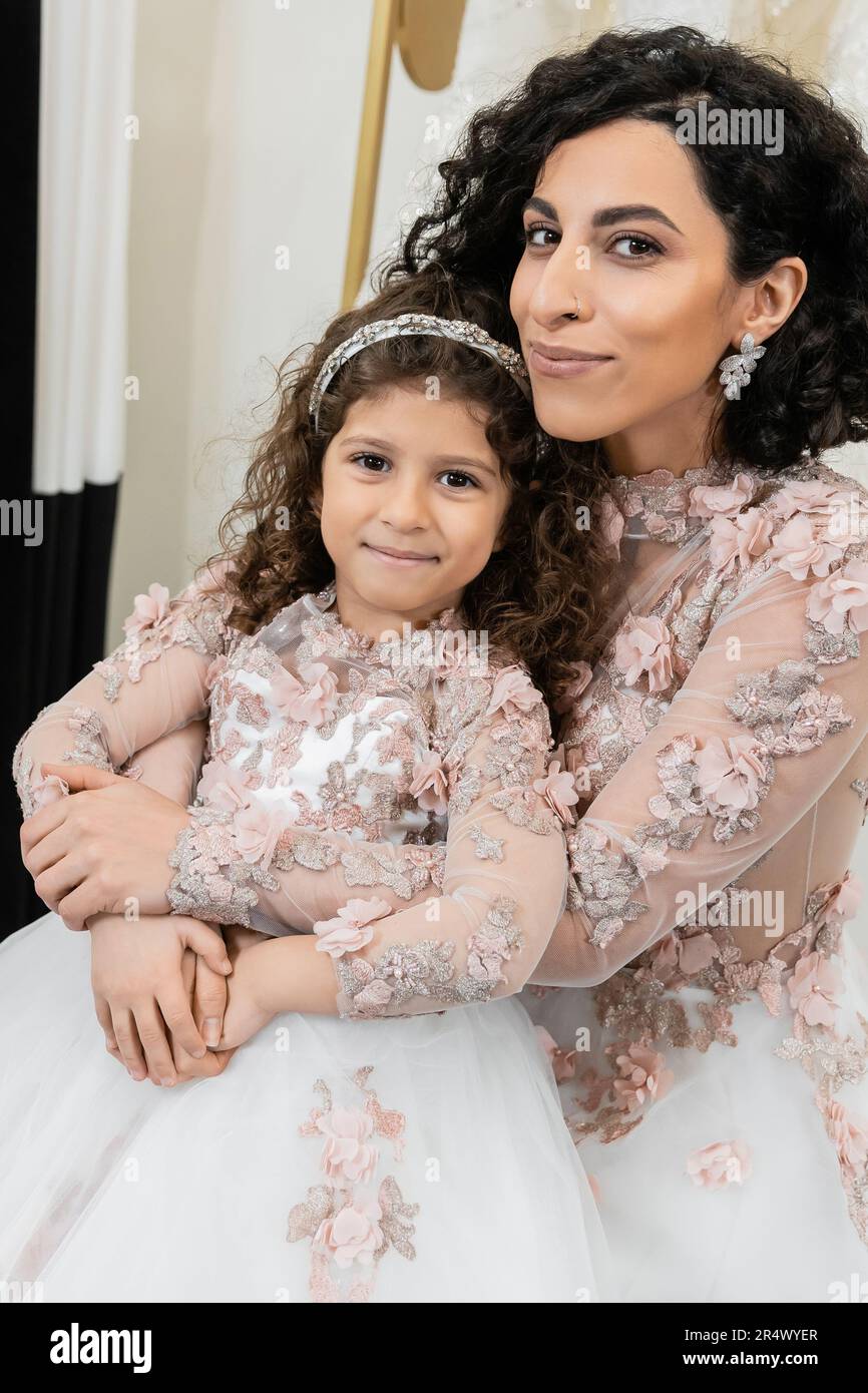 cheerful middle eastern bride in wedding dress hugging happy girl in ...