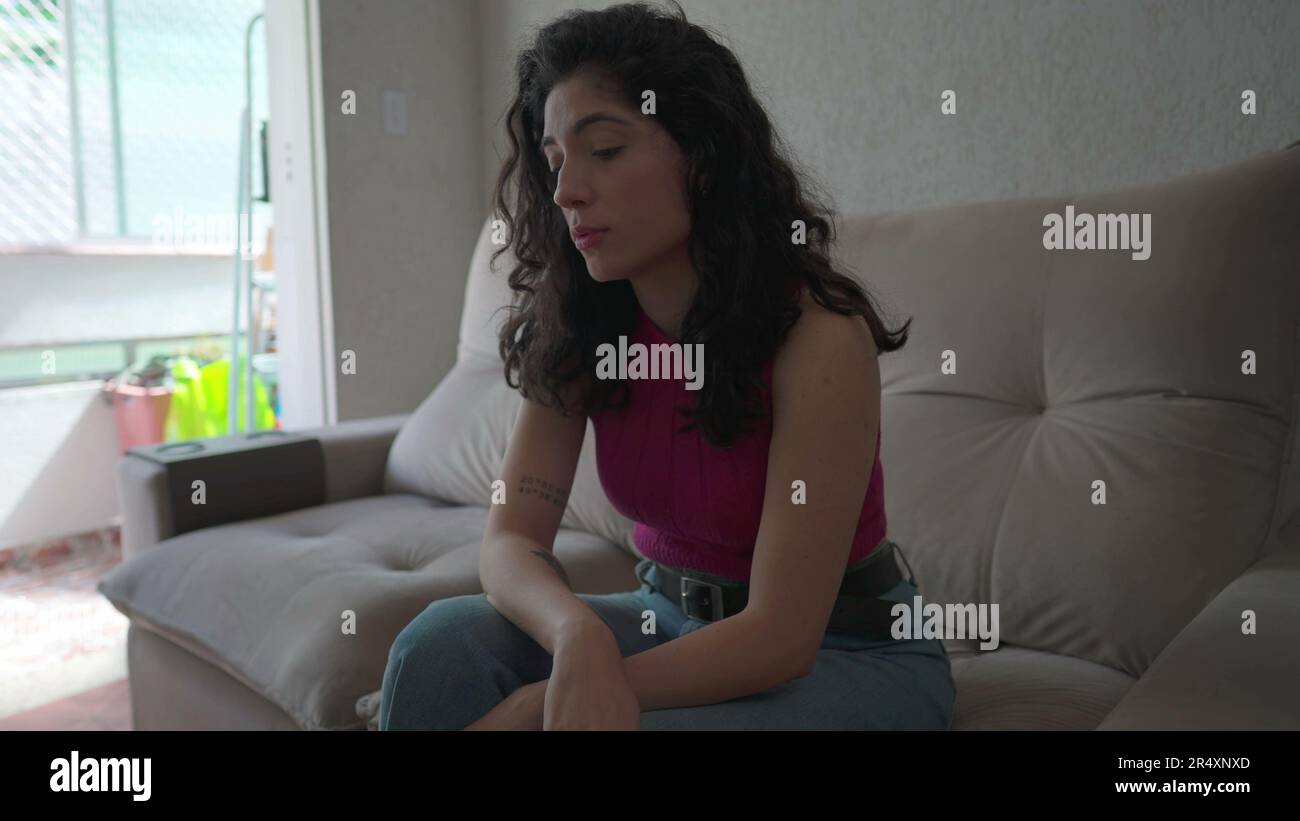 One pensive young woman sitting on couch at home with contemplative ...