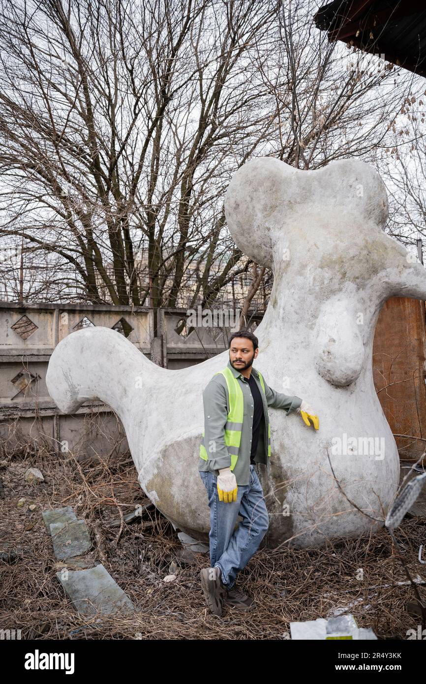 Indian worker in safety vest and gloves looking away while standing ...