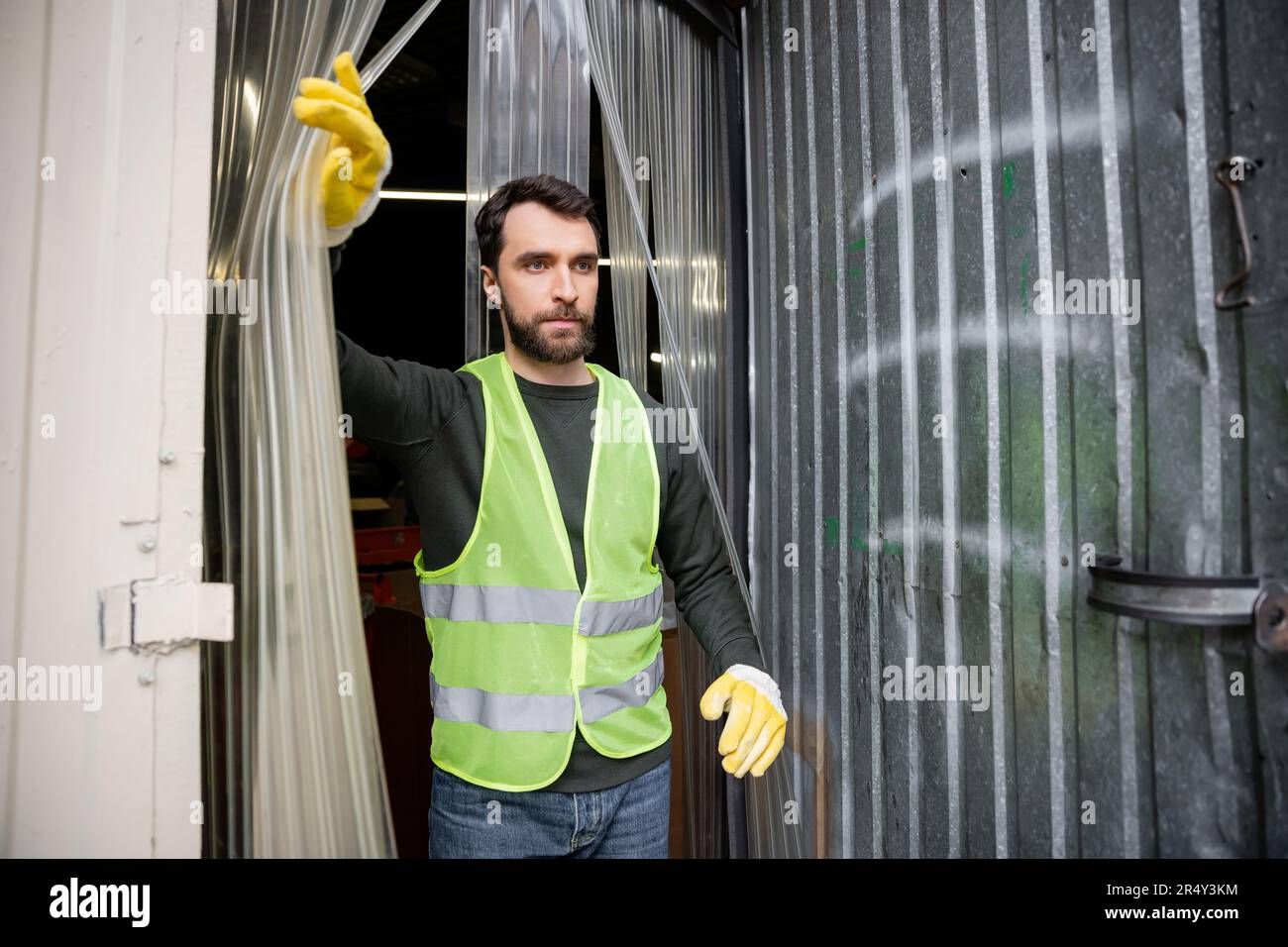 Bearded male worker in protective vest and gloves looking away and ...
