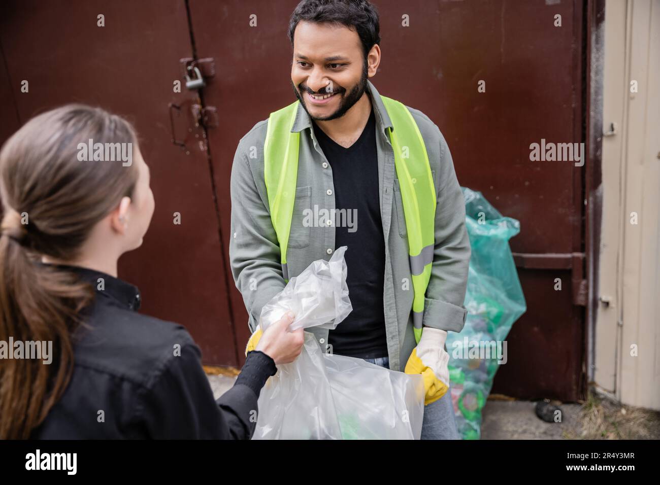 Smiling indian worker in safety vest and gloves taking trash bag from ...