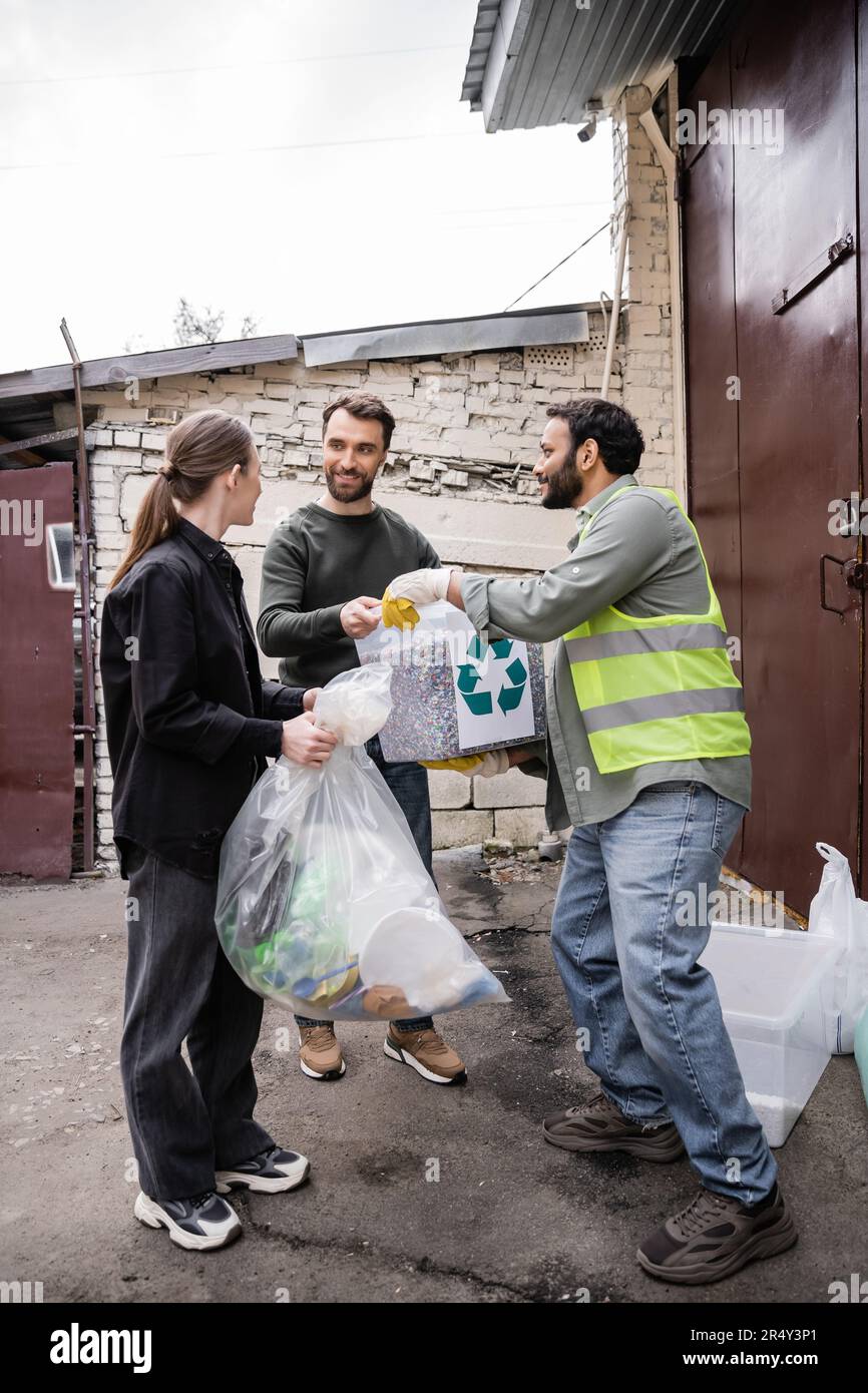 Smiling indian worker in protective vest taking trash from volunteers ...