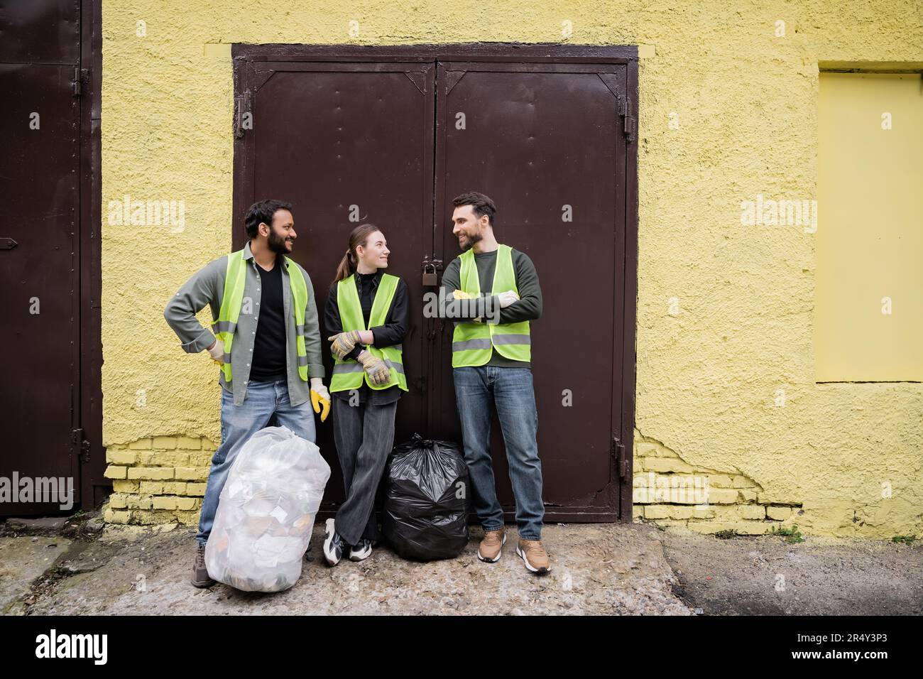 Smiling multiethnic workers in protective vests and gloves talking near ...