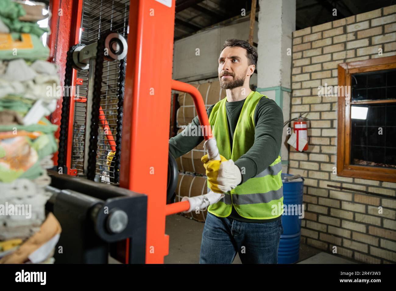 Bearded worker in fluorescent vest and gloves using hand pallet truck ...