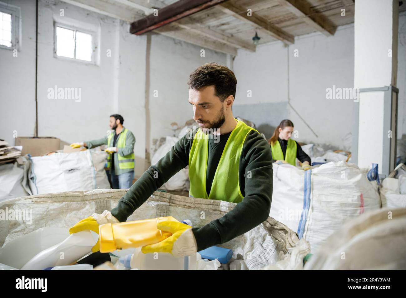 Bearded worker in protective gloves and vest taking plastic trash from ...
