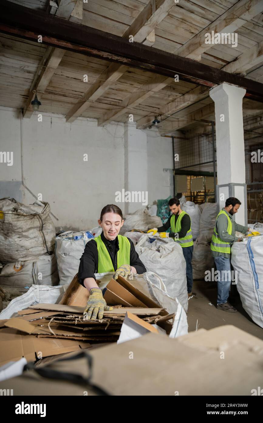 Positive worker in gloves and vest putting cardboard in sack while ...