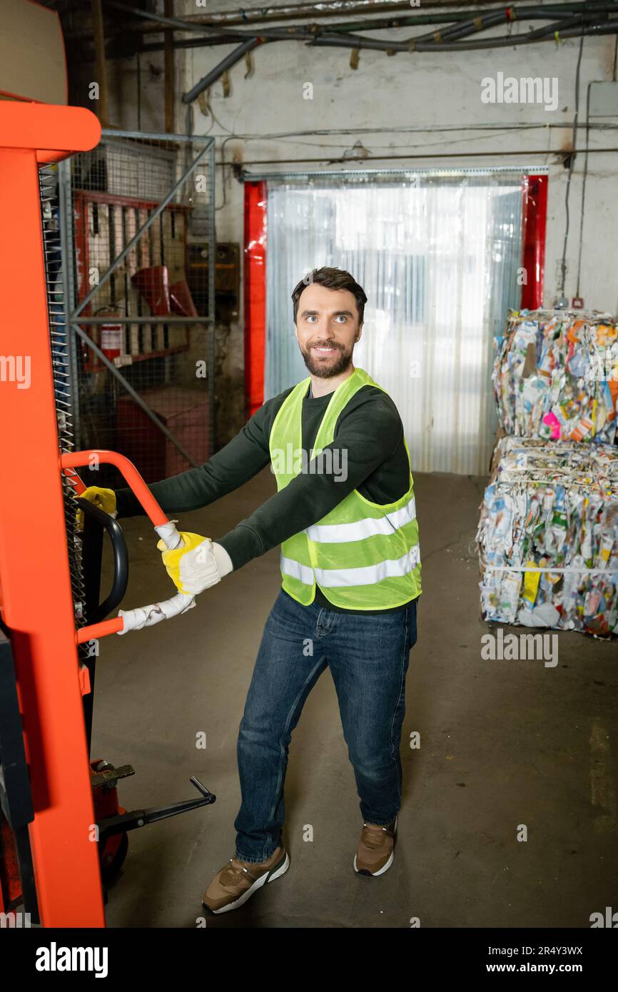 Smiling worker in safety vest and gloves pulling hand pallet truck ...