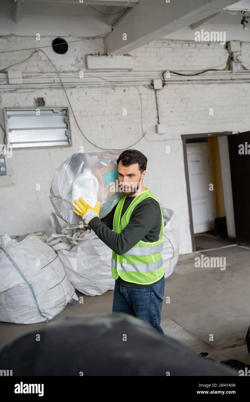 Bearded worker in protective vest and gloves carrying plastic bag with ...