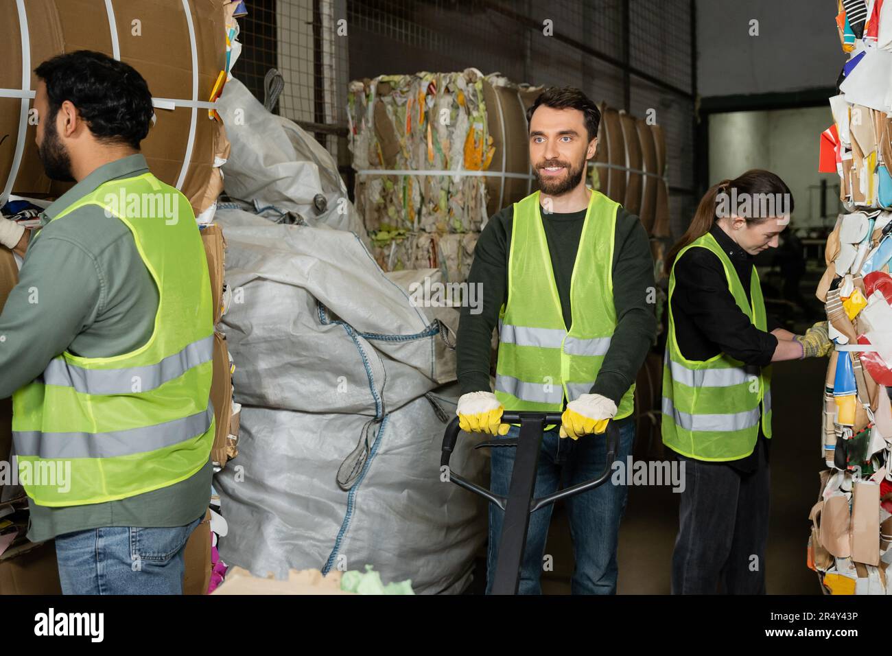 Smiling worker in protective vest and gloves standing near hand pallet ...