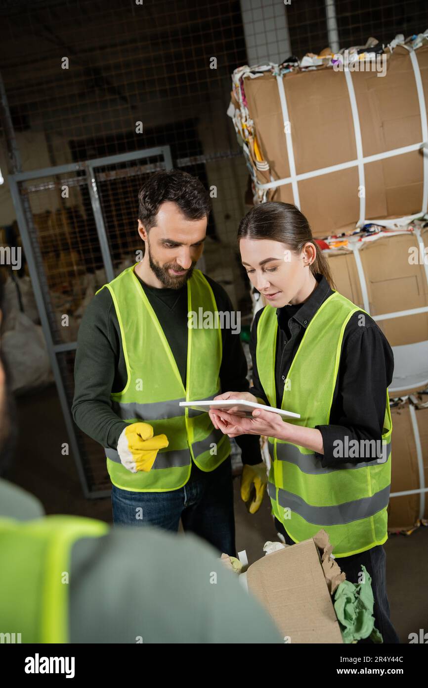 Male worker in safety vest and gloves talking to young colleague using ...