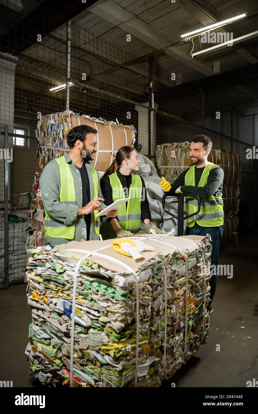 Cheerful sorter in protective vest and gloves talking to interracial ...