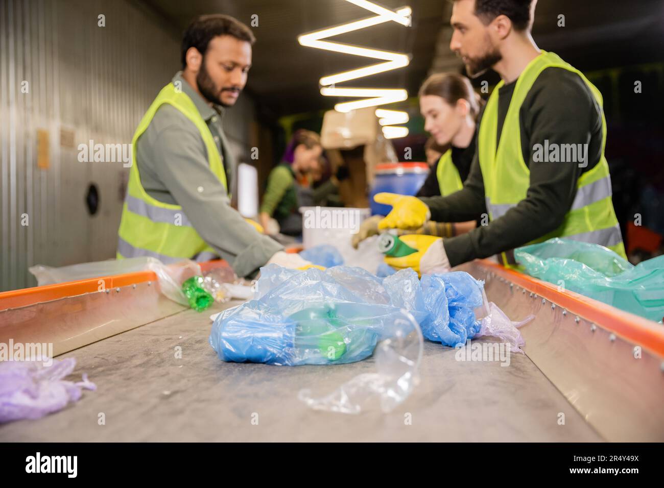 Plastic trash on conveyor near blurred multiethnic workers in high visibility vests and gloves ...