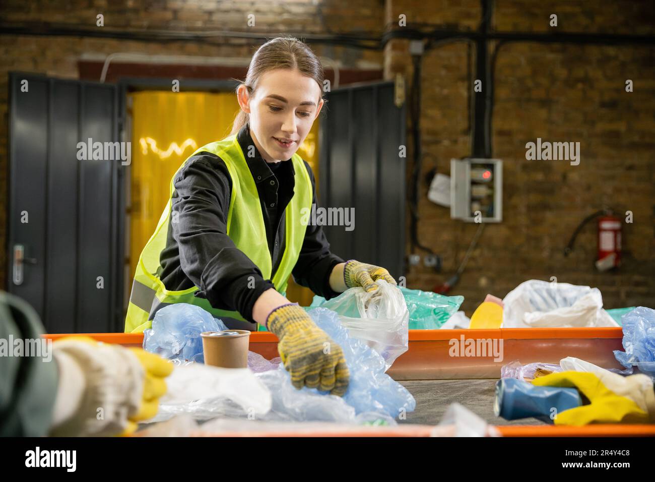 Smiling young worker in protective vest and gloves sorting garbage near ...