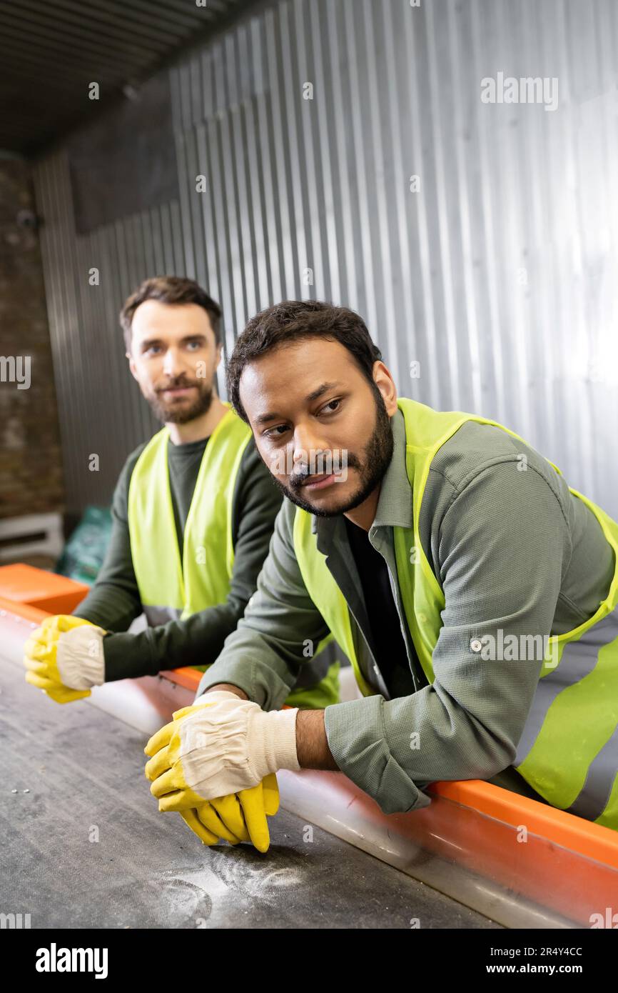 Indian sorter in protective vest and gloves looking away near conveyor ...