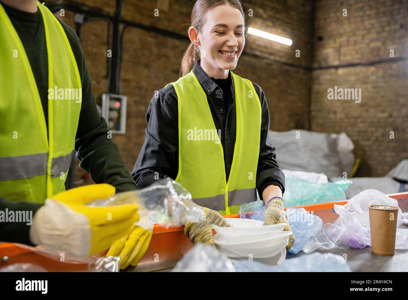 Positive young worker in gloves and protective vest holding plastic ...