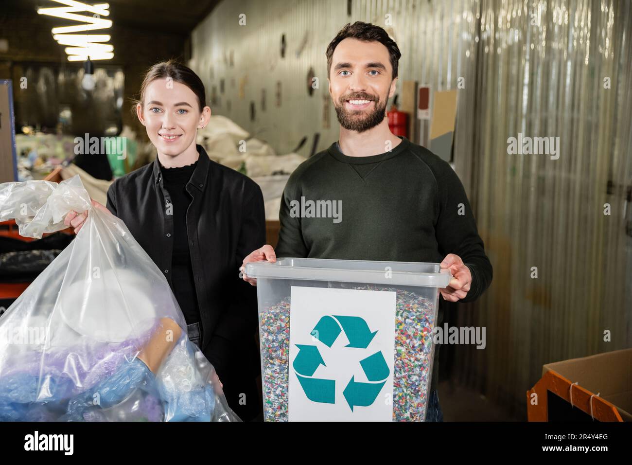 Cheerful volunteers looking at camera while holding trash bin and bag ...