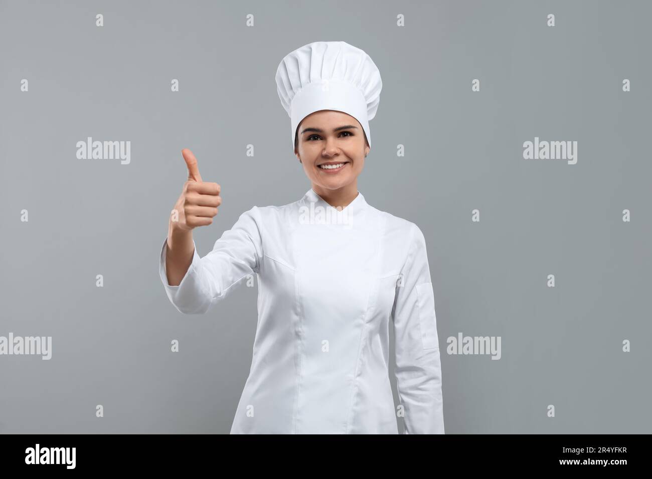 Happy female chef wearing uniform and cap showing thumbs up on light ...