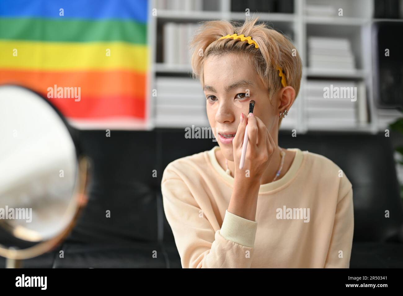 A young Asian queer man looking in the mirror and applying foundation ...