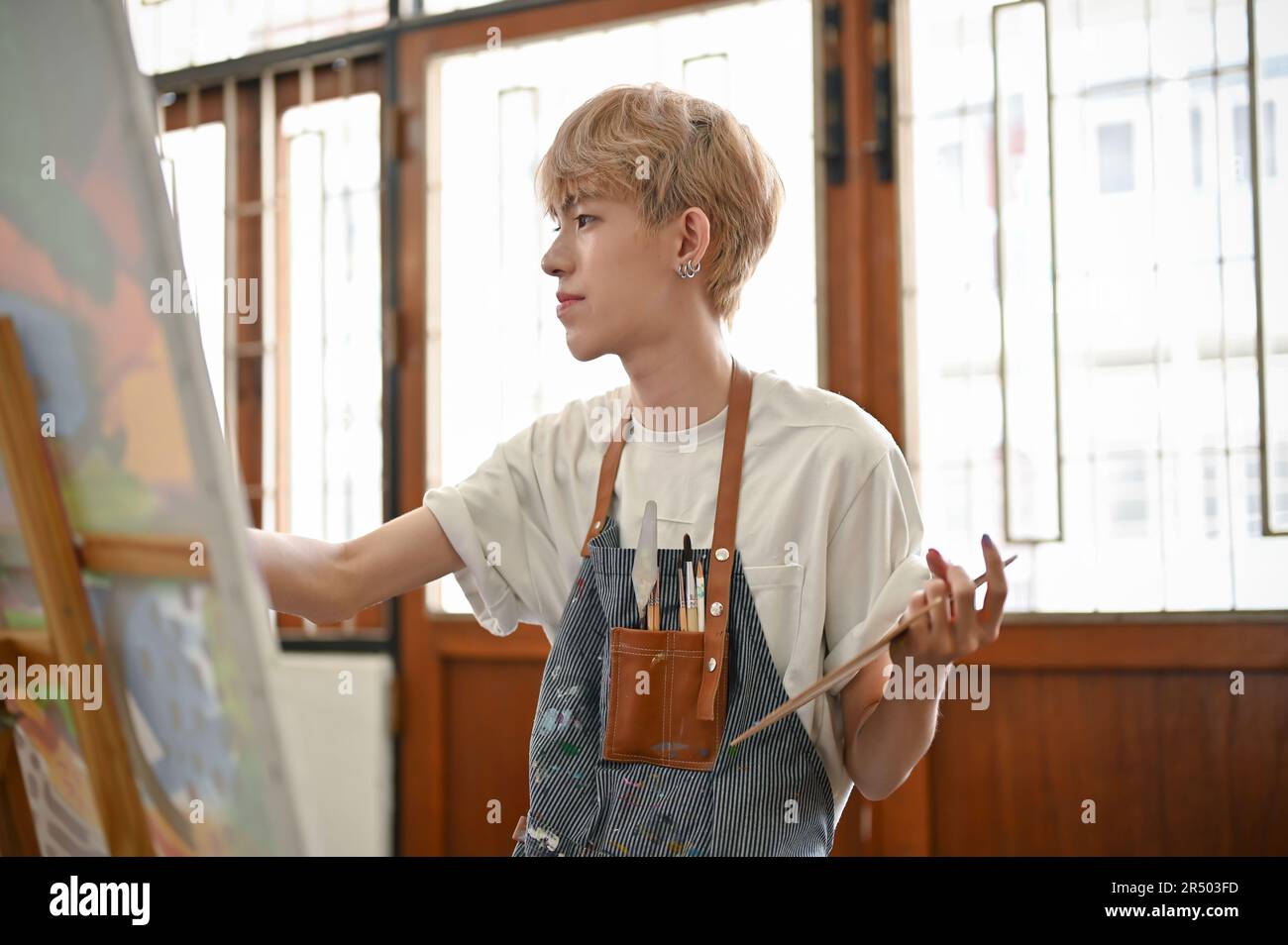 An attractive and creative young Asian queer artist in an apron focuses ...