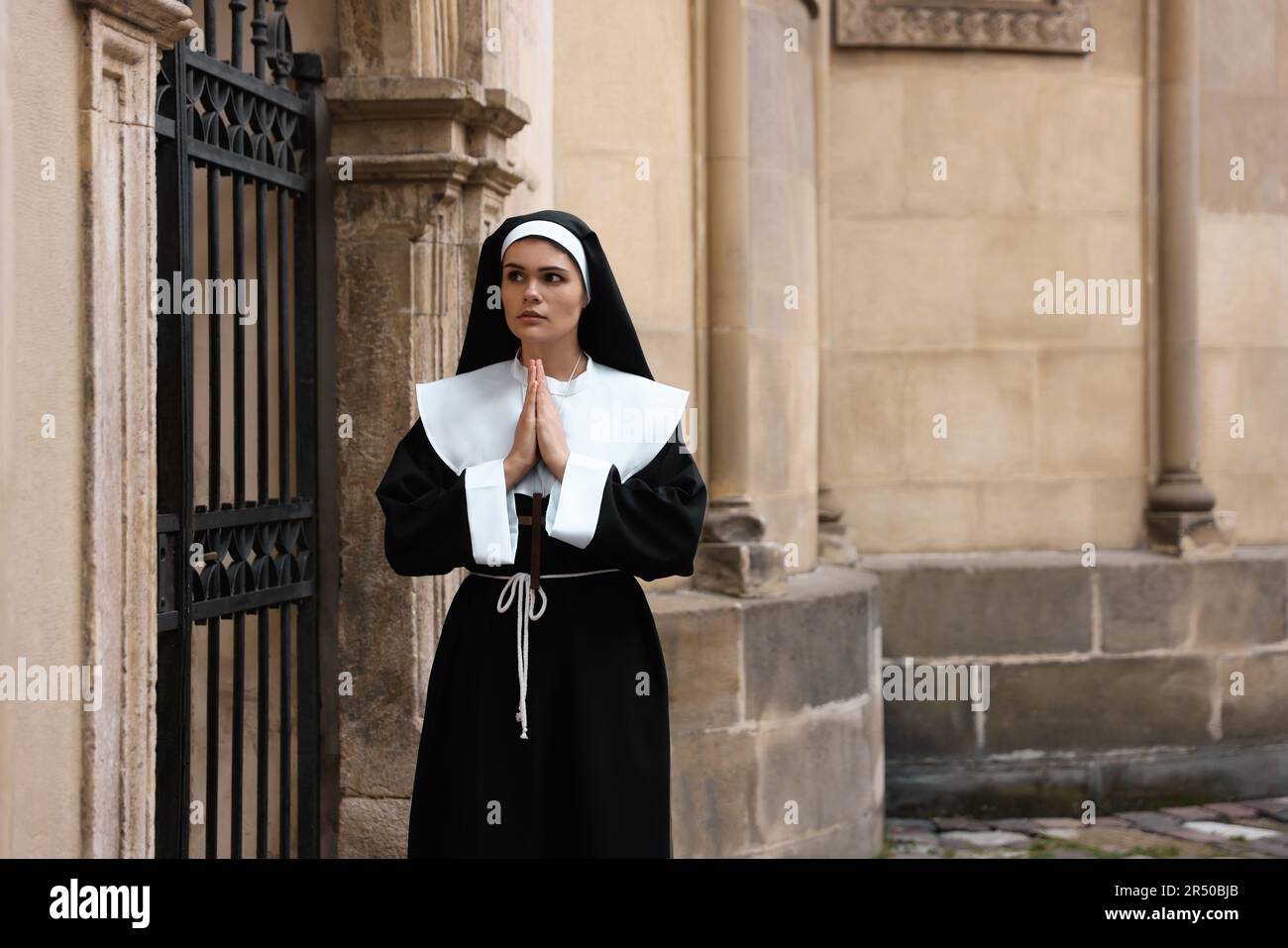 Young nun with hands clasped together praying near metal door outdoors ...