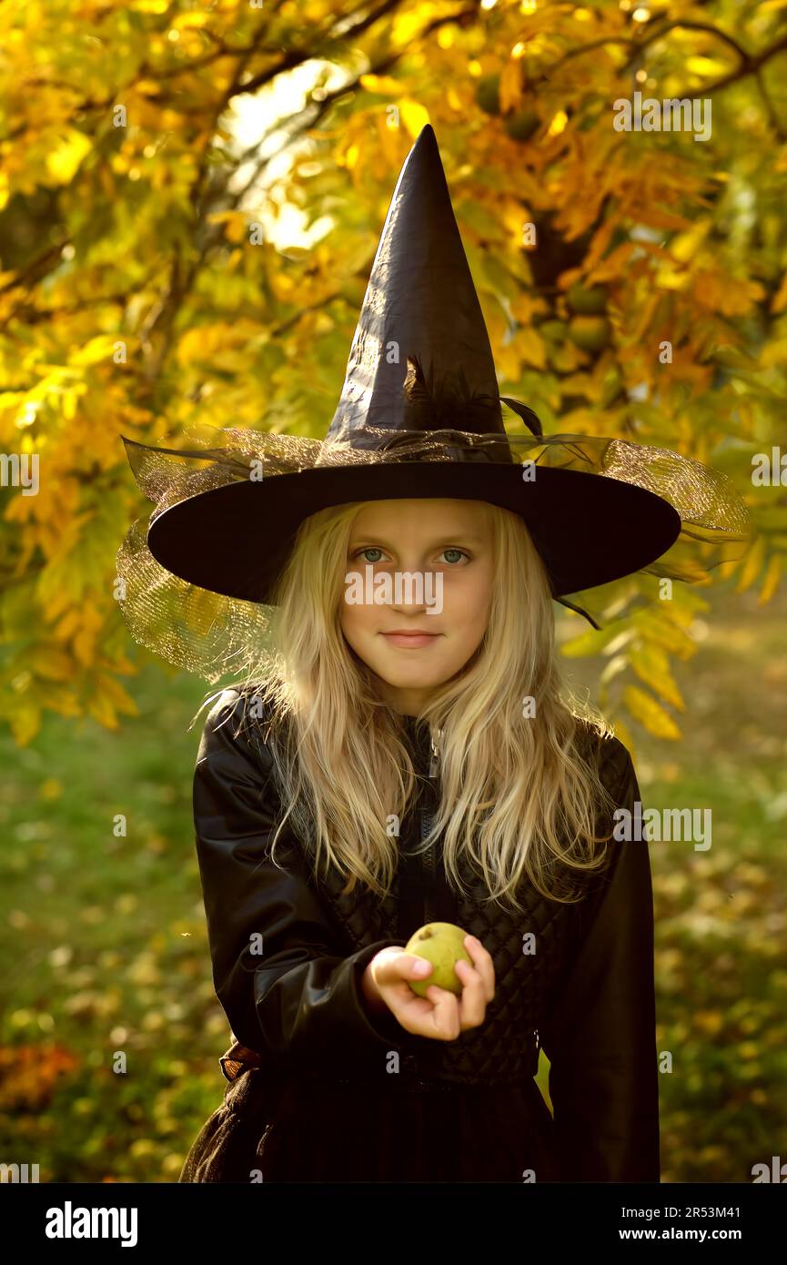 A little girl in a witch costume holds out an apple, looking at the ...