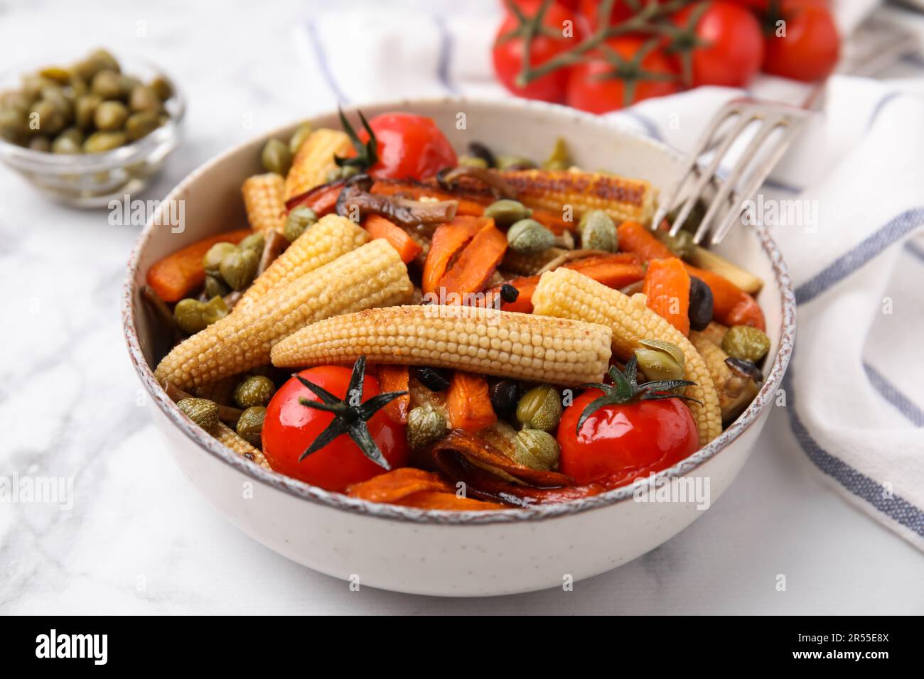 Tasty roasted baby corn with tomatoes and capers on white marble table ...