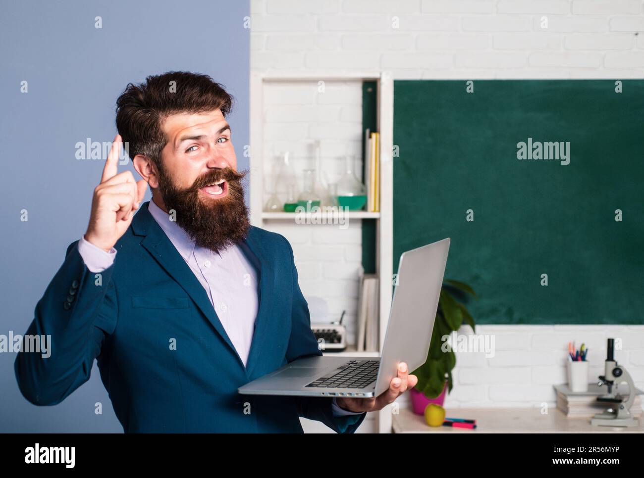 Excited teacher with pointing finger hold laptop notebook in classroom ...