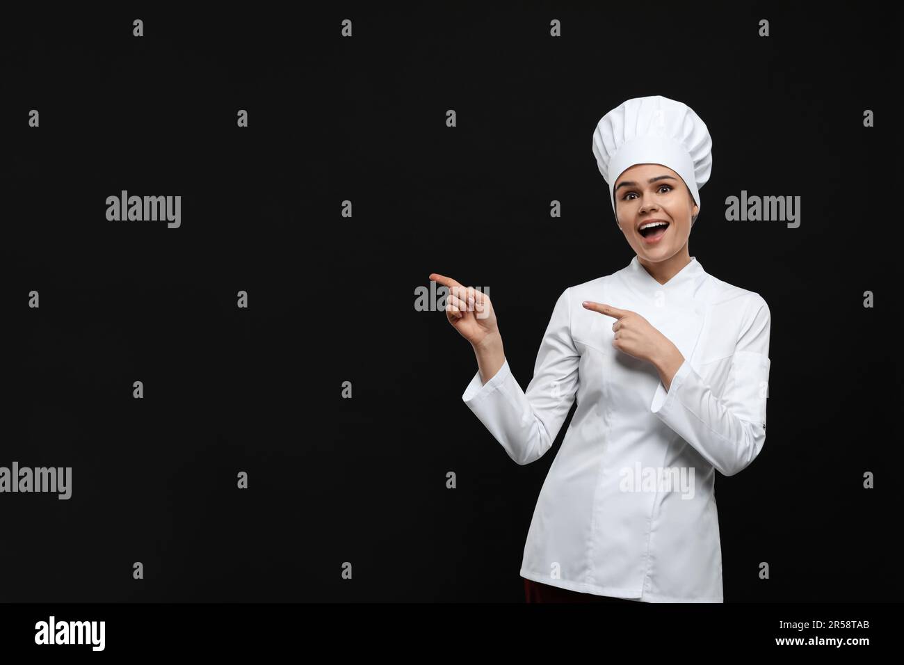 Happy female chef pointing at something on black background. Space for ...
