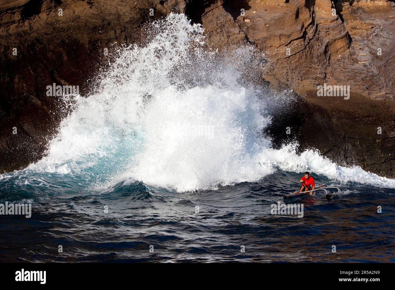One man paddling an outrigger canoe next to big cliffs and crashing ...