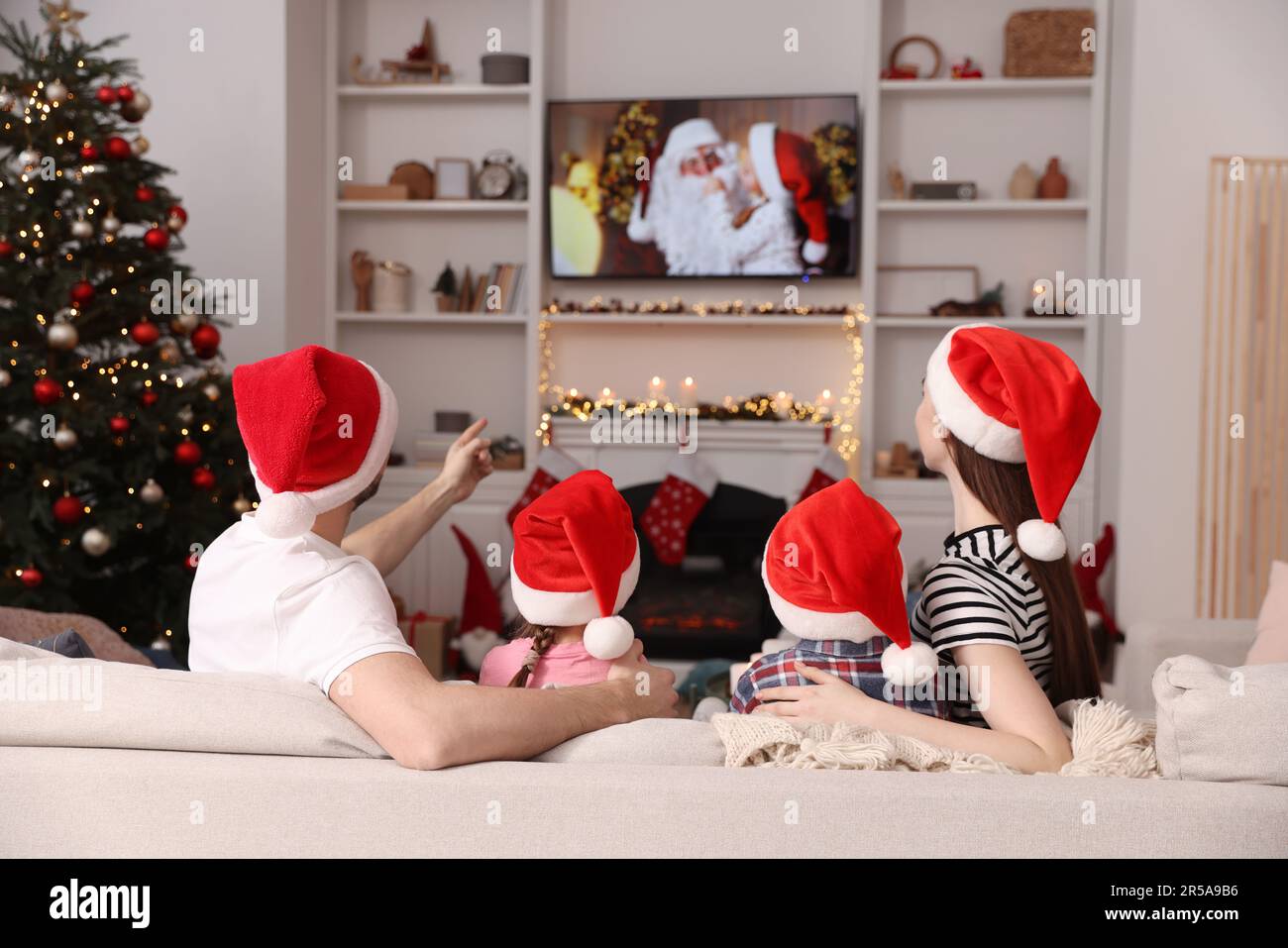 Family wearing Christmas hats watching TV in cosy room, back view. Winter holidays atmosphere ...