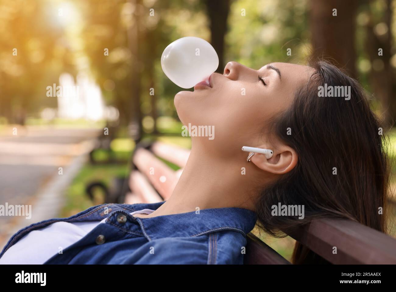 Beautiful young woman with wireless headphones blowing chewing gum on wooden bench outdoors ...