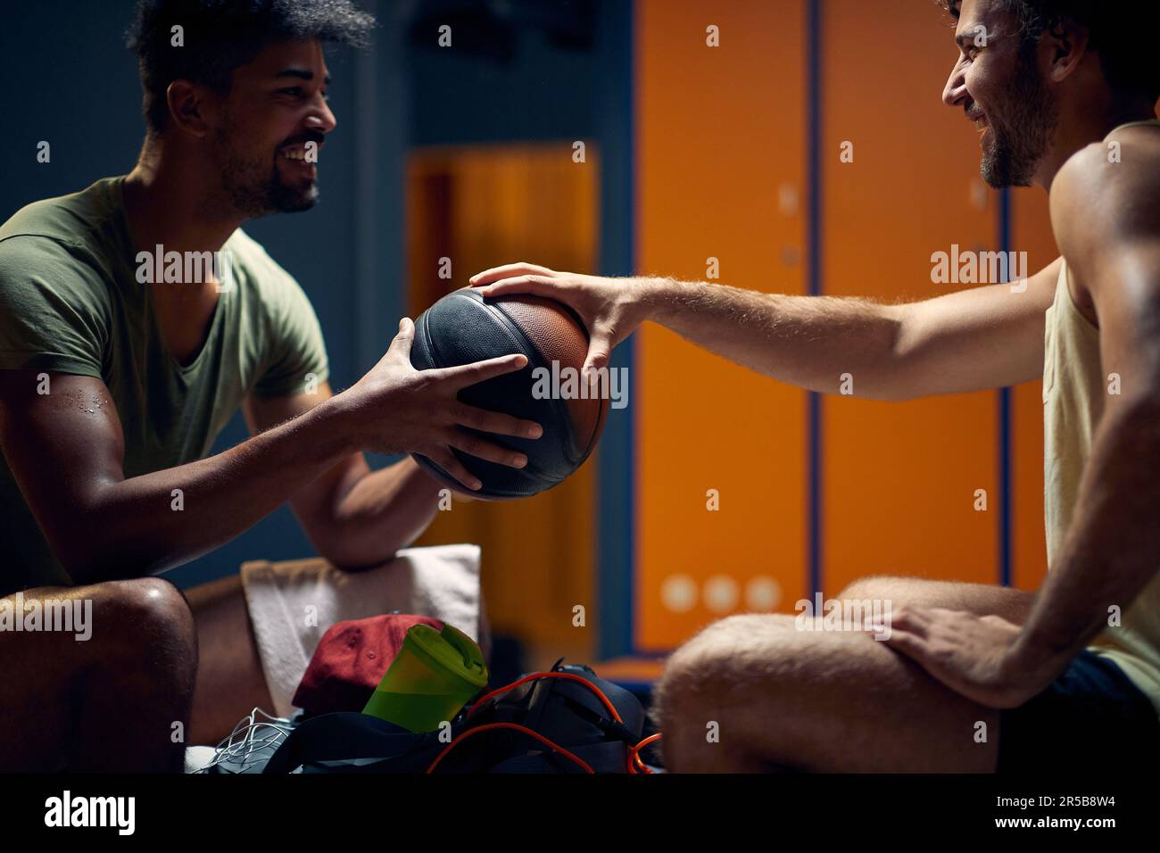 Joyful young men sitting face to face in gym locker room, holding ...