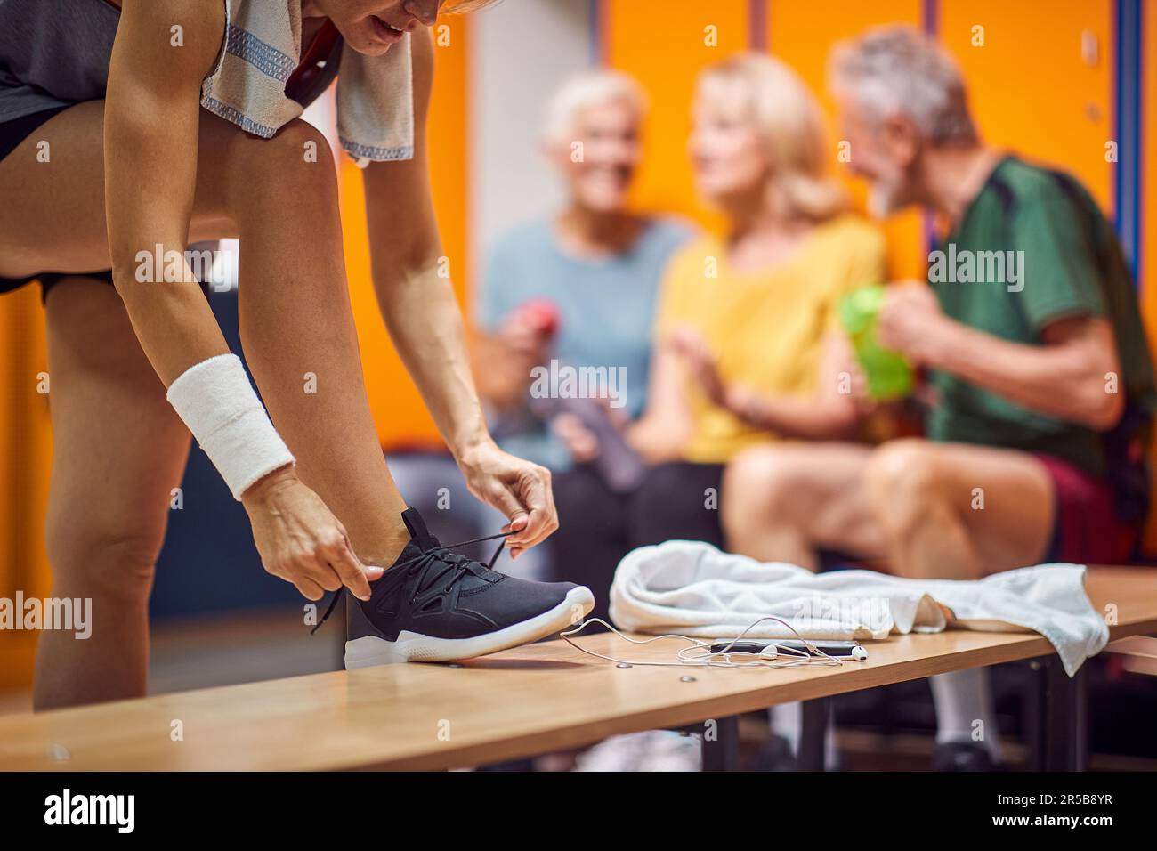 People in gym locker room. Young woman tying her shoe, seniors in the ...