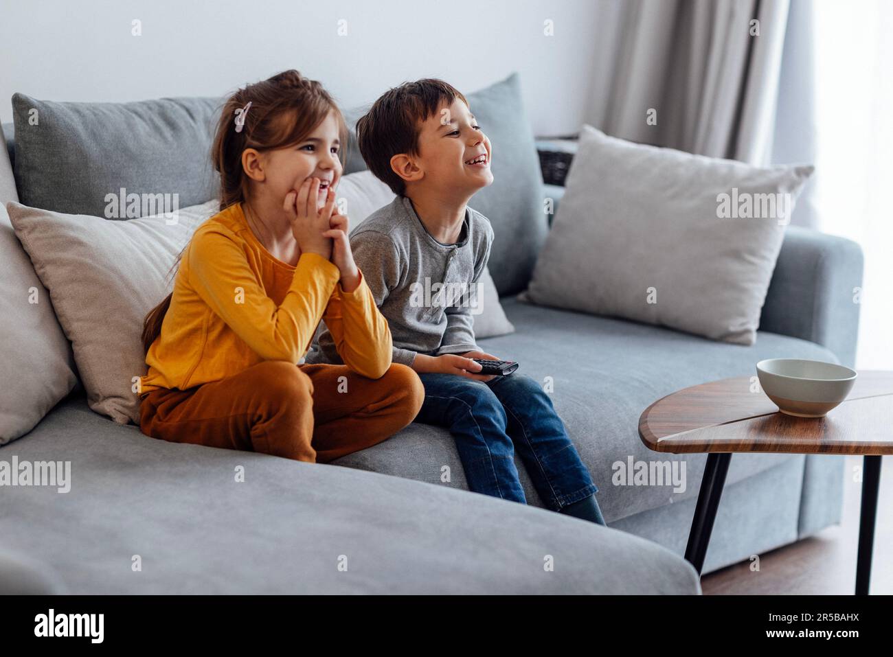 Two little kids are sitting on grey sofa and watching television. Cute ...
