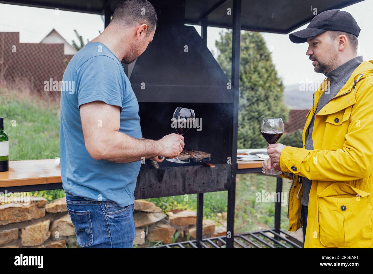 Two young men in casual clothes are roasting meat and drinking wine ...