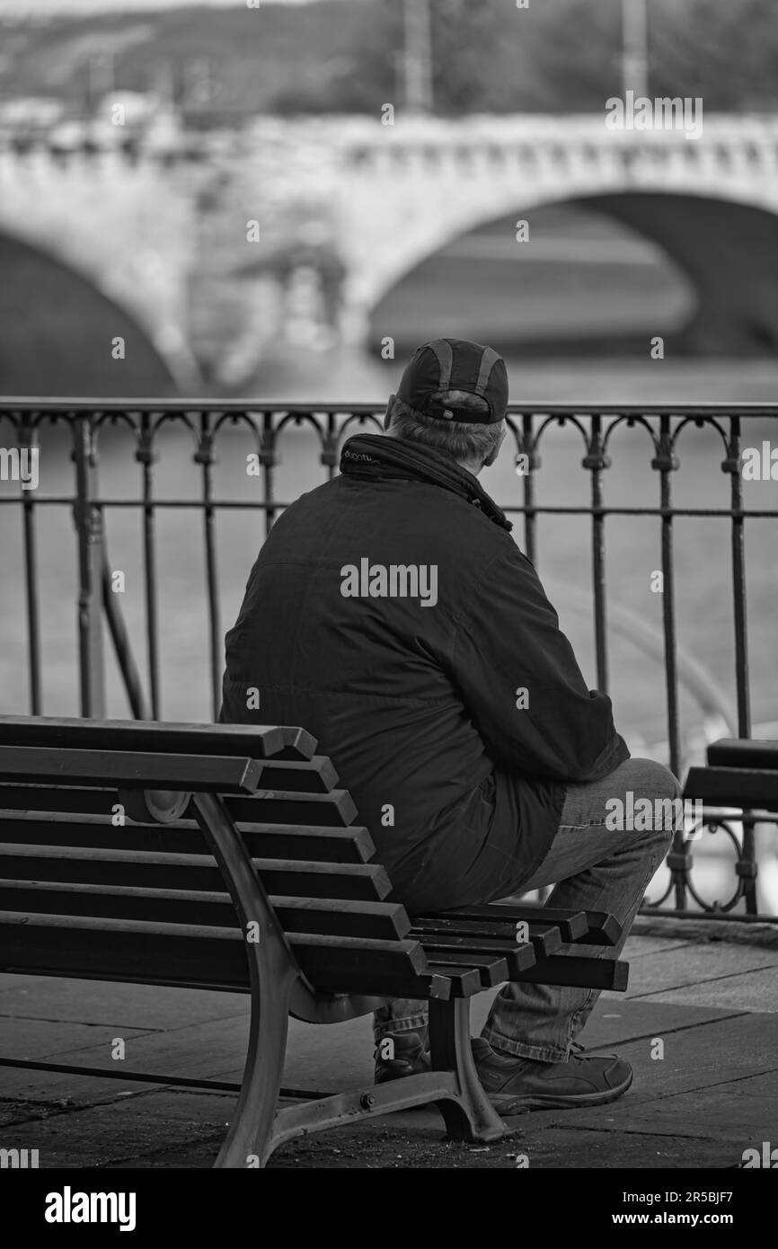 A grayscale of a man sitting alone on a park bench with a stunning ...
