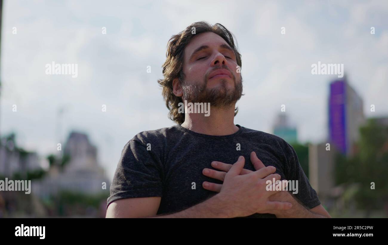 Tranquil young man putting hand in chest taking a deep breath outside ...